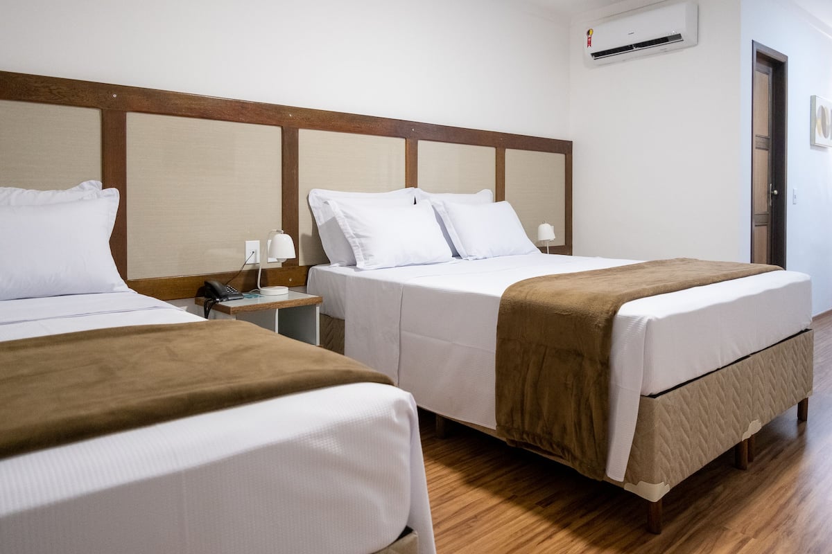 Quarto de hotel com duas camas, cabeceira de madeira, mesa de cabeceira com abajur e ar condicionado na parede.