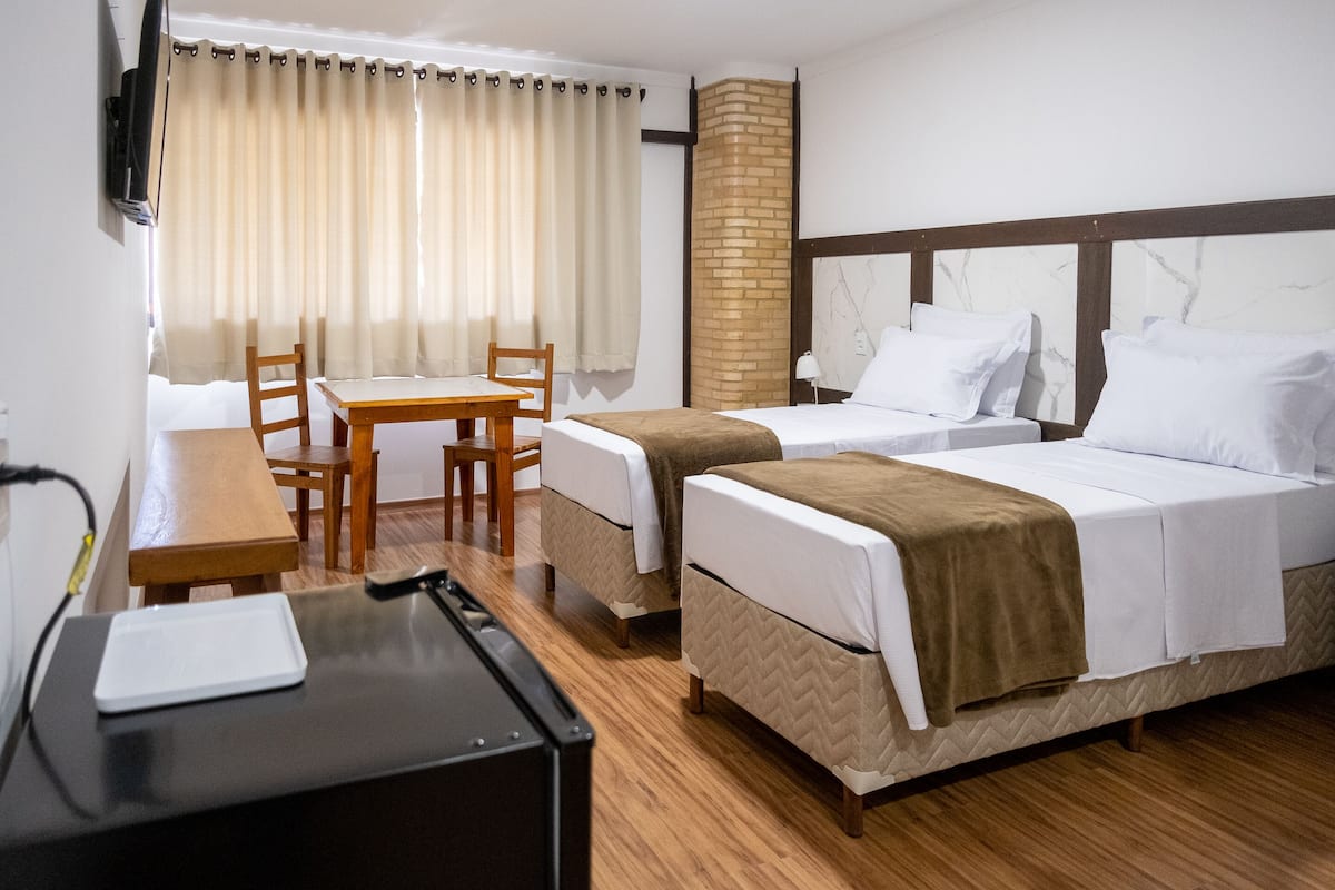 Quarto de hotel com duas camas, uma mesa de madeira, cadeiras, uma TV e uma parede de destaque em tijolo.