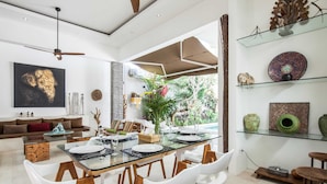 Uma área de refeições moderna com mesa de vidro, cadeiras brancas e vista para um jardim.