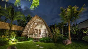 Uma casa com telhado de palha e varanda, rodeada por vegetação exuberante e palmeiras.