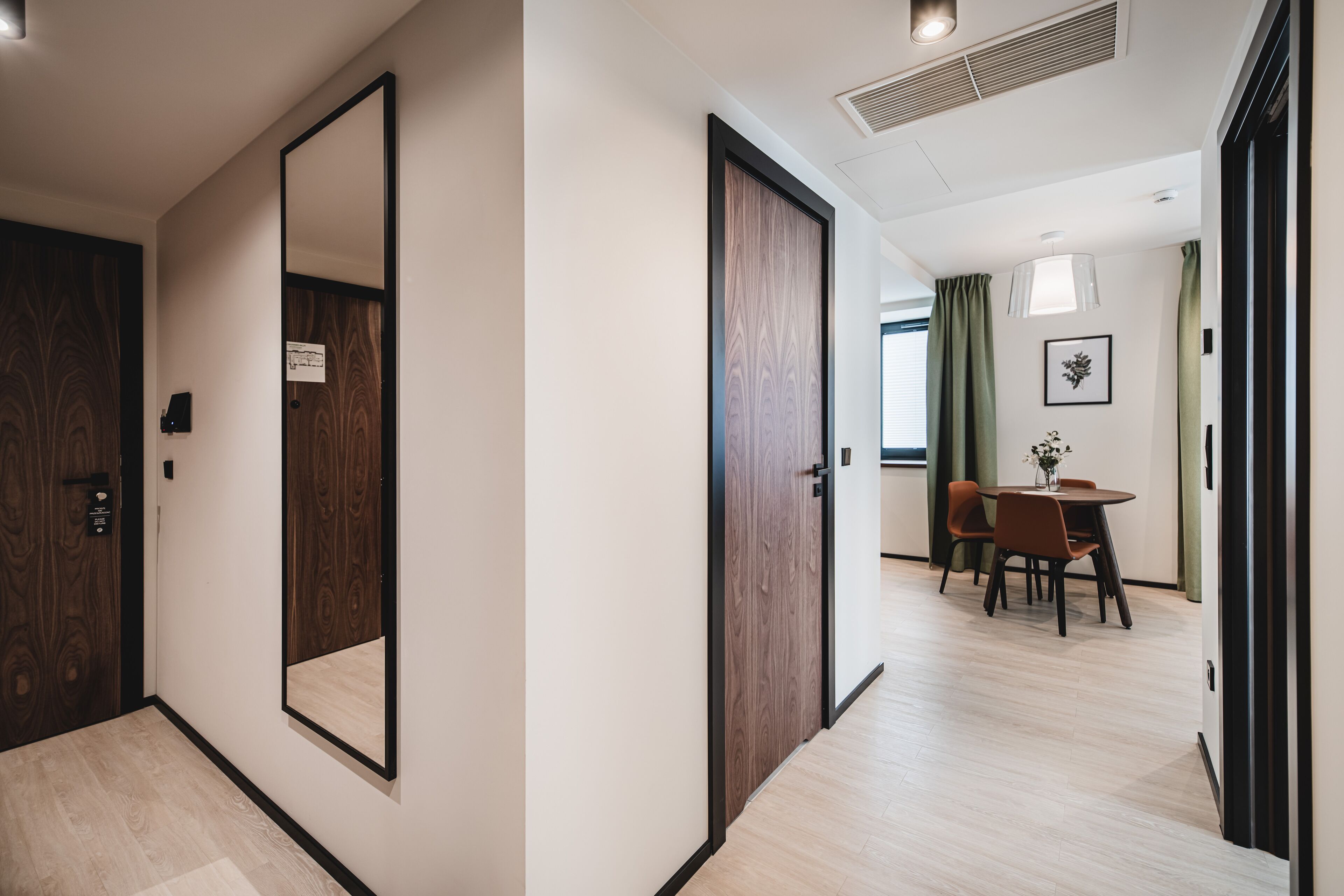 Um corredor com porta de madeira e espelho, que dá para uma área de refeições com mesa e cadeiras.