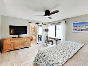 Um quarto com cama, televisão, cômoda, escrivaninha e ventilador de teto.
