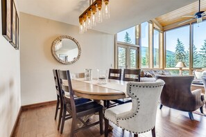 Uma sala de jantar com uma mesa redonda, cadeiras, um lustre e uma janela grande com vista para árvores.