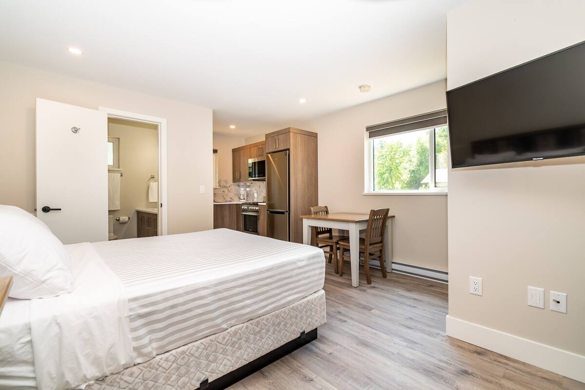 Quarto com cama, pequena cozinha, área de refeições e TV na parede.
