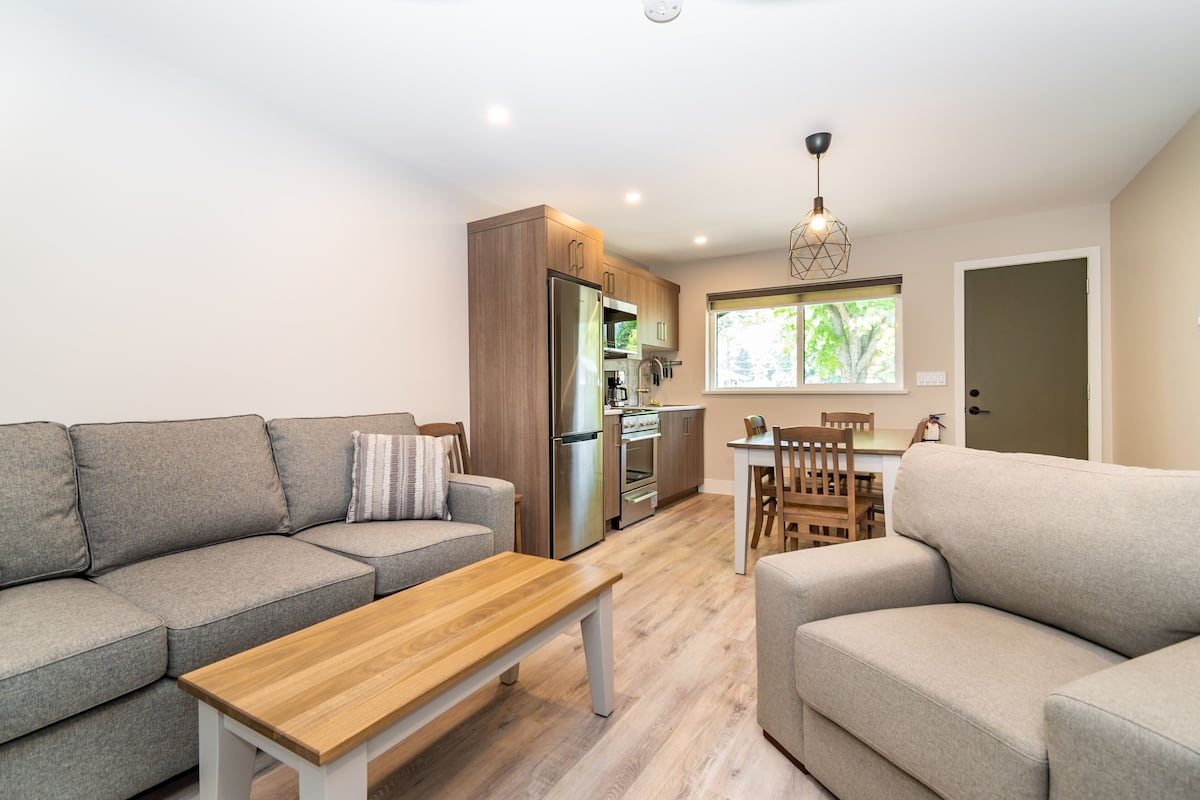 Uma sala de estar moderna com um sofá cinzento, uma mesa de centro de madeira e uma área de cozinha com frigorífico, forno e mesa de jantar.