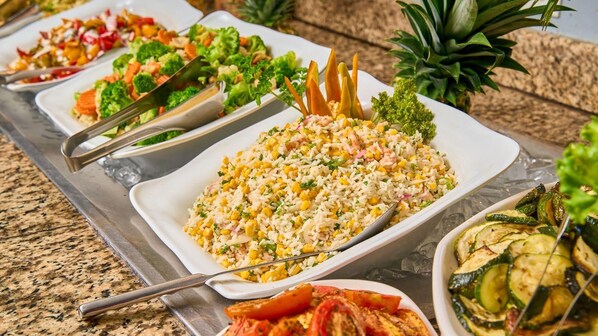 Mesa de buffet com diversos pratos, incluindo arroz, legumes e comidas grelhadas.
