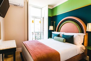 Quarto de hotel com cama, escrivaninha, televisão e janela com vista para o exterior.