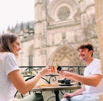 Duas pessoas sentadas à mesa ao ar livre, com um edifício histórico ao fundo.