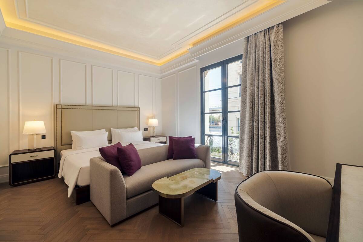 Quarto de hotel com cama, sofá, poltrona, mesa pequena e mesa de cabeceira com luminárias.