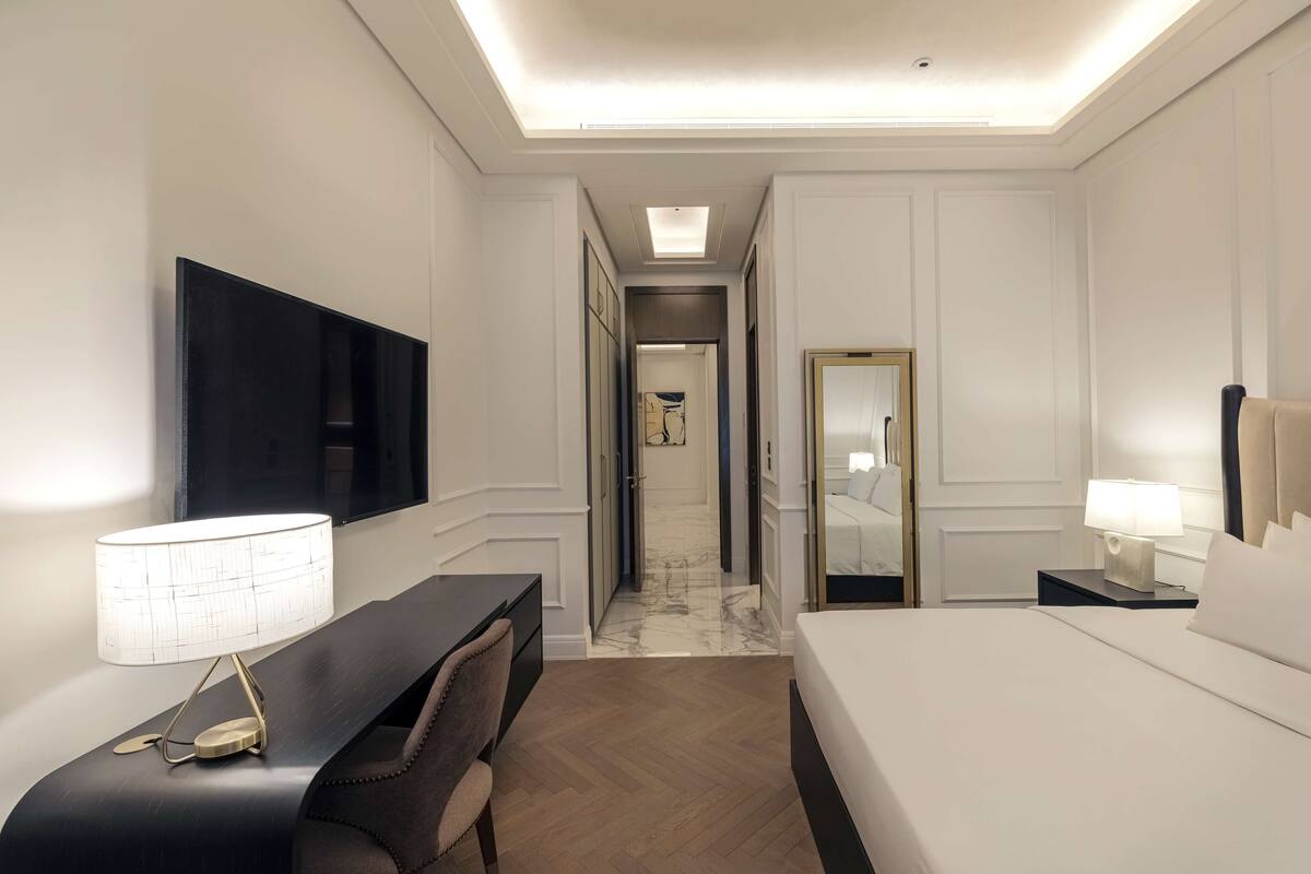 Quarto de hotel com cama, escrivaninha com luminária, cadeira, televisão e espelho.