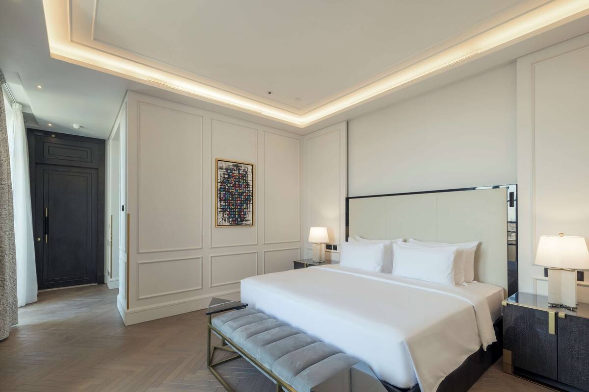 Quarto de hotel com uma cama grande, mesas de cabeceira, um banco e uma obra de arte na parede.