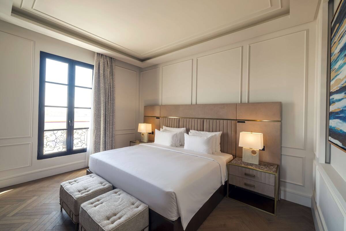 Quarto de hotel com uma cama grande, duas mesas de cabeceira com abajures, uma janela com cortinas e um quadro na parede.