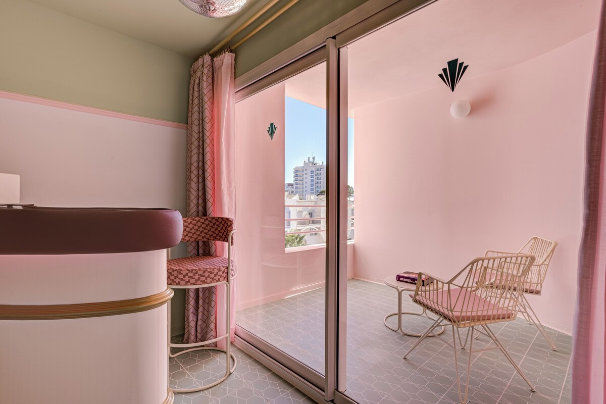 Uma varanda com decoração rosa, uma cadeira estofada, uma mesa pequena e vista para edifícios.