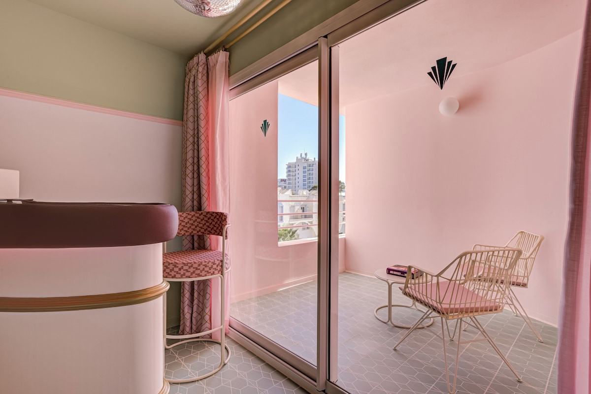 Uma varanda com decoração rosa, uma cadeira estofada, uma mesa pequena e vista para edifícios.