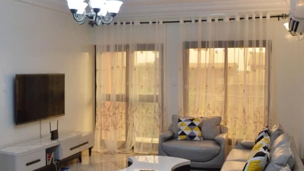 Uma sala de estar moderna com televisão de tela plana, um sofá cinza com almofadas amarelas, uma mesa de centro e um lustre.