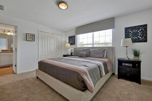 Quarto com uma cama grande, janela com persianas, mesa de cabeceira com luminária e uma obra de arte na parede.