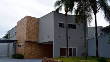 Um edifício moderno de dois andares com parte em tijolo e fachada cinzenta, apresentando entrada com portão e um pequeno jardim com palmeiras.