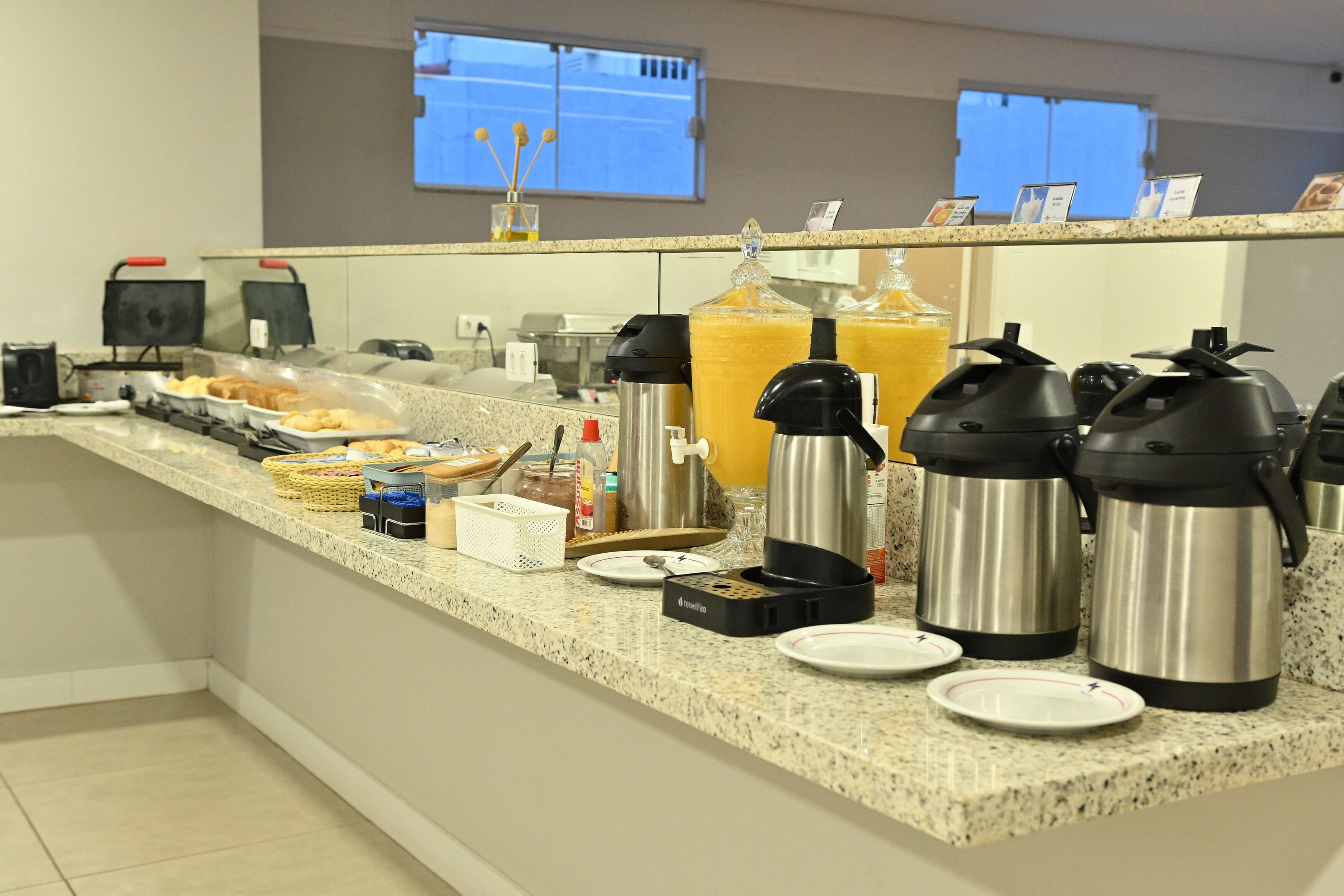 Uma mesa de buffet com diversos alimentos, máquinas de café e um dispensador de suco.