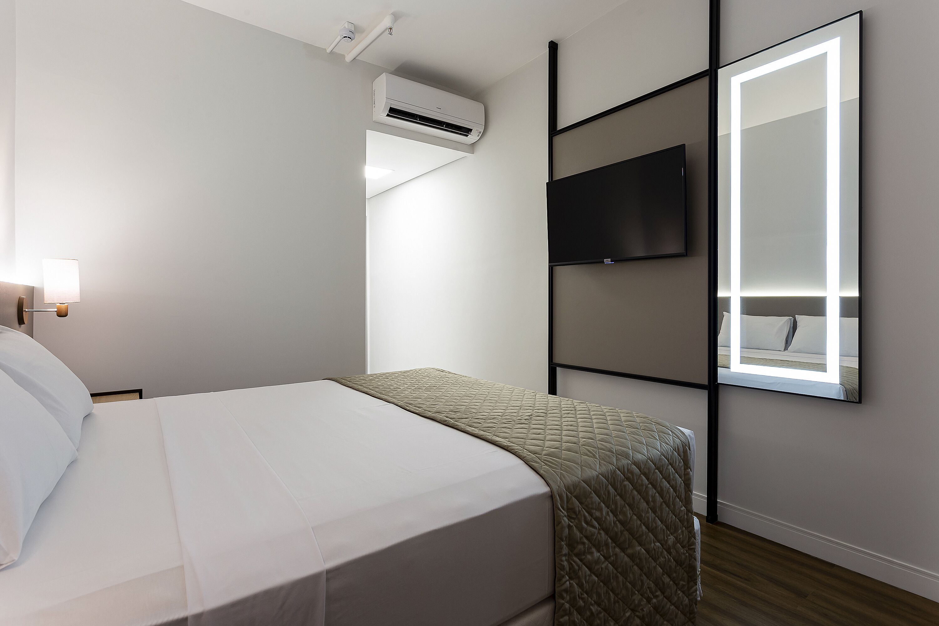 Quarto de hotel com cama, unidade de ar condicionado, TV de tela plana na parede e um espelho grande refletindo parte do quarto.