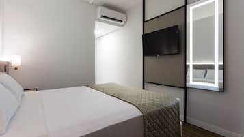 Quarto de hotel com cama, unidade de ar condicionado, TV de tela plana na parede e um espelho grande refletindo parte do quarto.