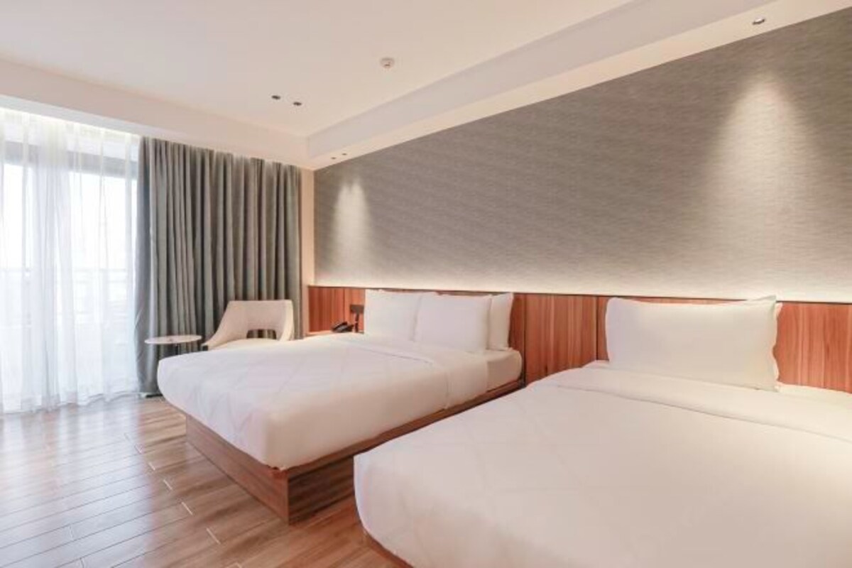 Quarto de hotel moderno com duas camas, piso de madeira, uma janela grande com cortinas e cabeceira fixada na parede.