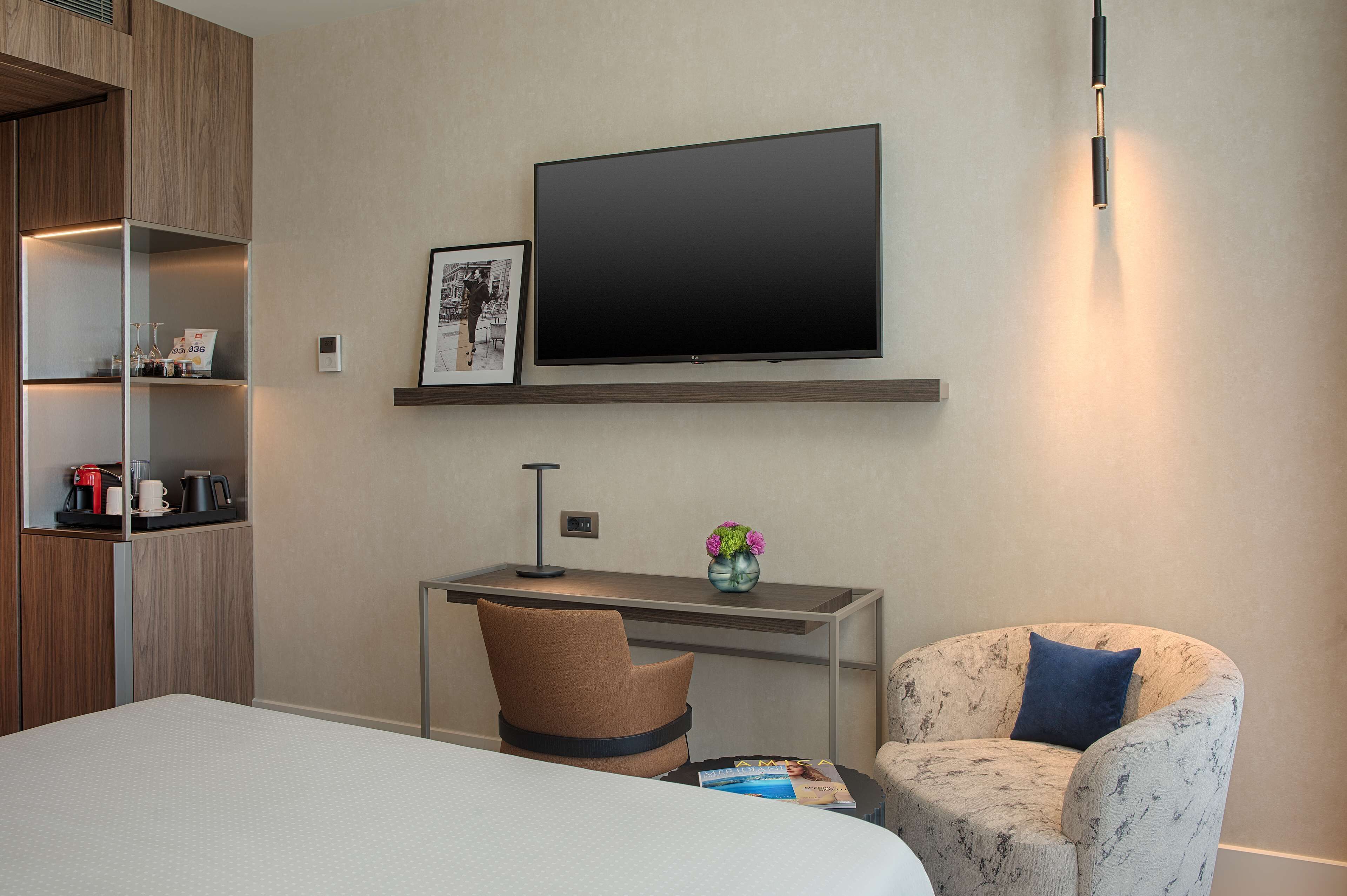 Quarto de hotel com cama, escrivaninha com televisão de tela plana, cadeira, pequena mesa com um vaso de flores e prateleira com objetos.