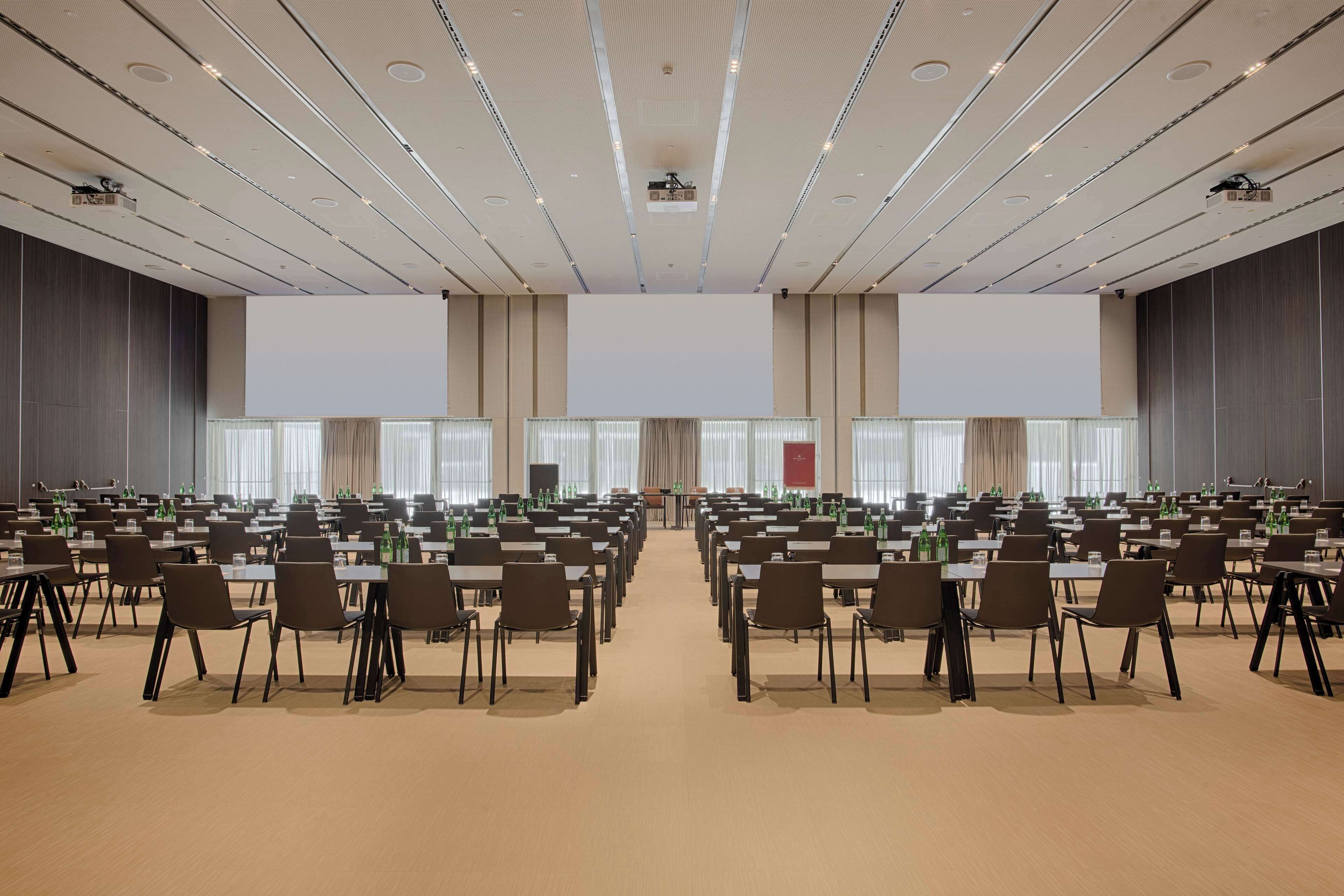 Uma grande sala de conferências com fileiras de mesas e cadeiras, um palco com um púlpito e uma tela grande na frente.