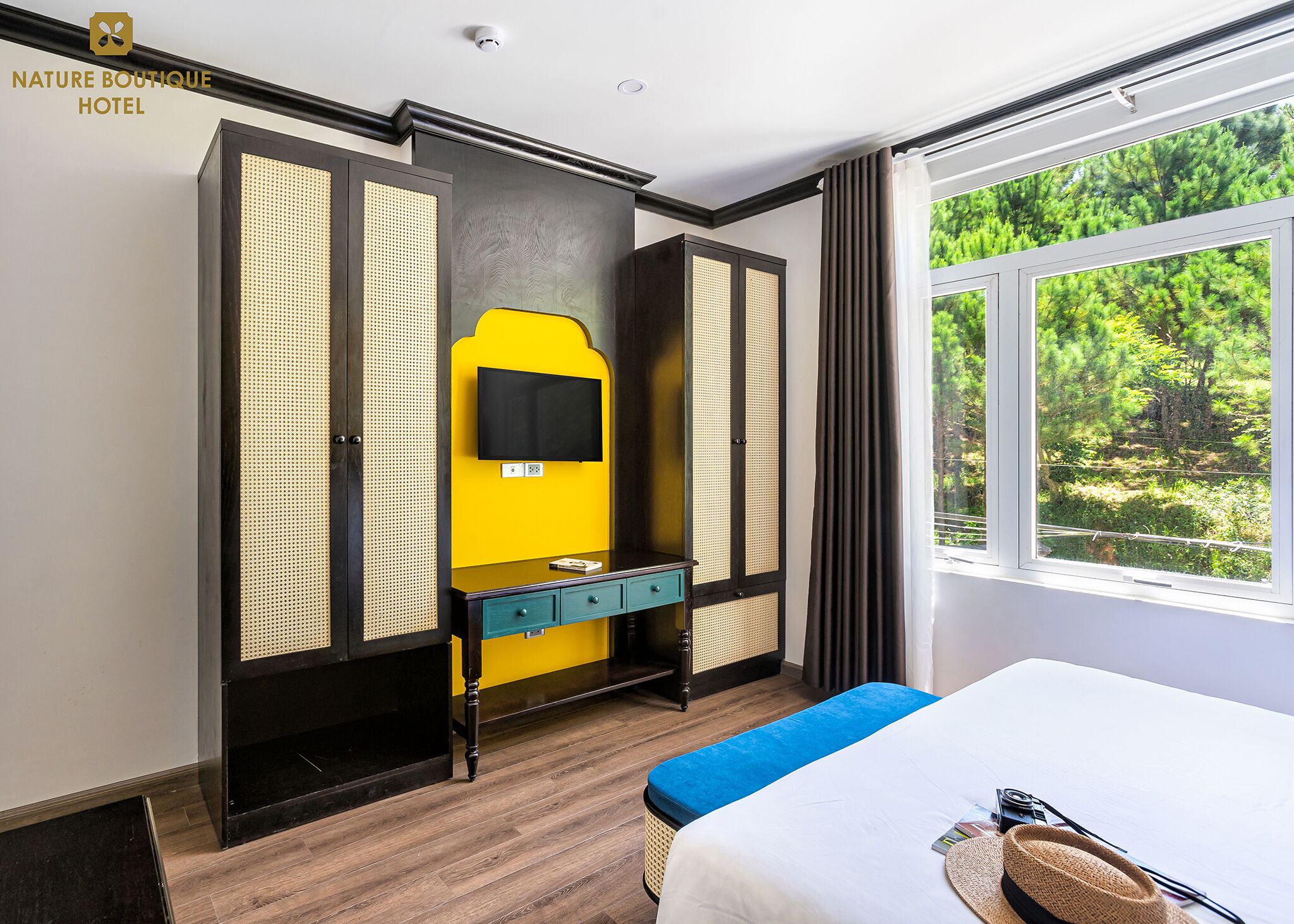 Quarto de hotel com uma cama, uma escrivaninha com televisão, guarda-roupa e janela com vista para árvores.