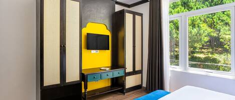 Quarto de hotel com uma cama, uma escrivaninha com televisão, guarda-roupa e janela com vista para árvores.