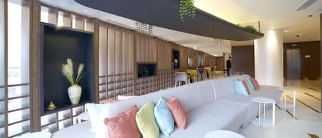 Um hall de hotel moderno com um sofá grande, pequenas mesas e uma tela de televisão.
