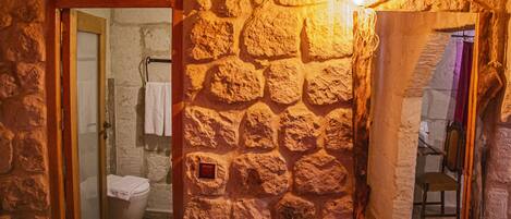 Casa de banho com paredes de pedra, um sanita, uma toalha pendurada na parede e uma porta de madeira que dá para outro quarto.