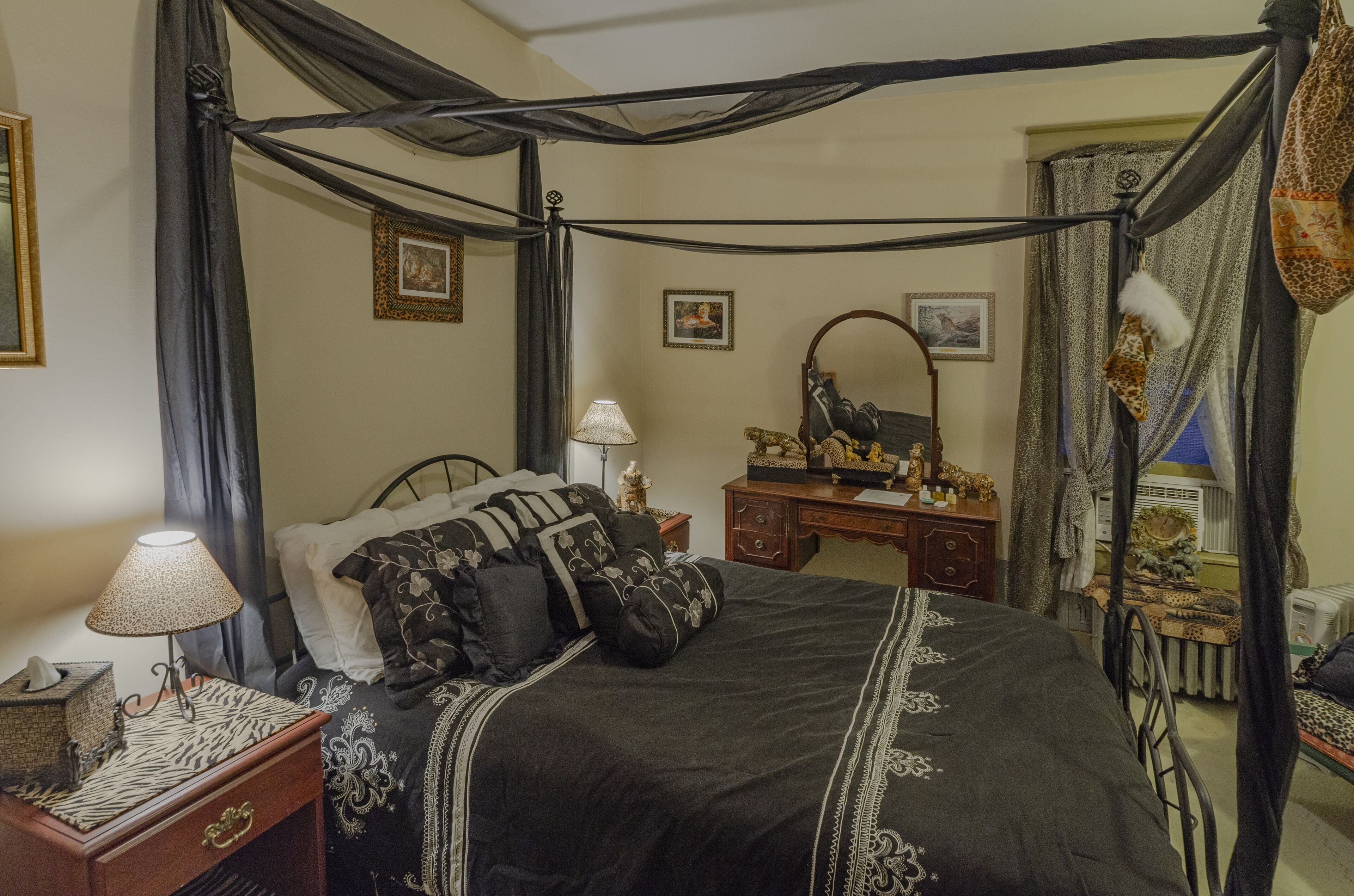 Uma cama de dossel com um edredom escuro e várias almofadas, uma mesa de cabeceira com abajur, um penteadeira com espelho e quadros nas paredes.