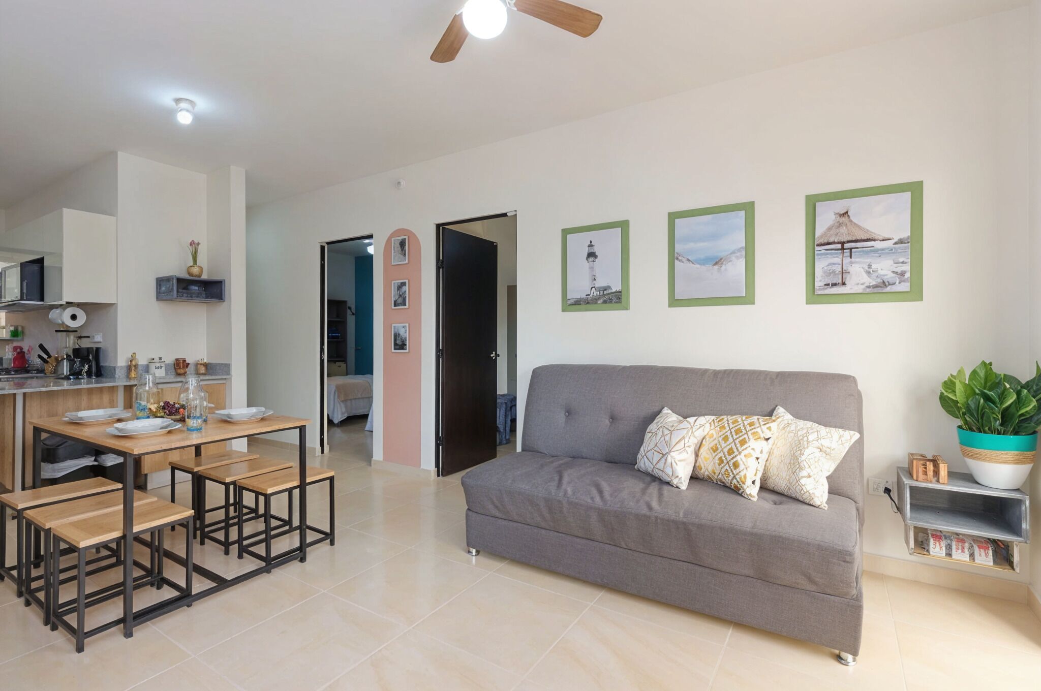 Uma sala de estar moderna com um sofá cinzento, mesa de jantar de madeira e cadeiras. Há quadros nas paredes, um ventilador de teto e um vaso de planta.