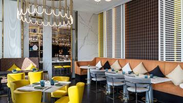 Interior moderno de restaurante com cadeiras amarelas, área de bar e espaço de estar com sofá e mesas.