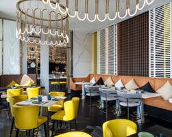 Interior moderno de restaurante com cadeiras amarelas, área de bar e espaço de estar com sofá e mesas.