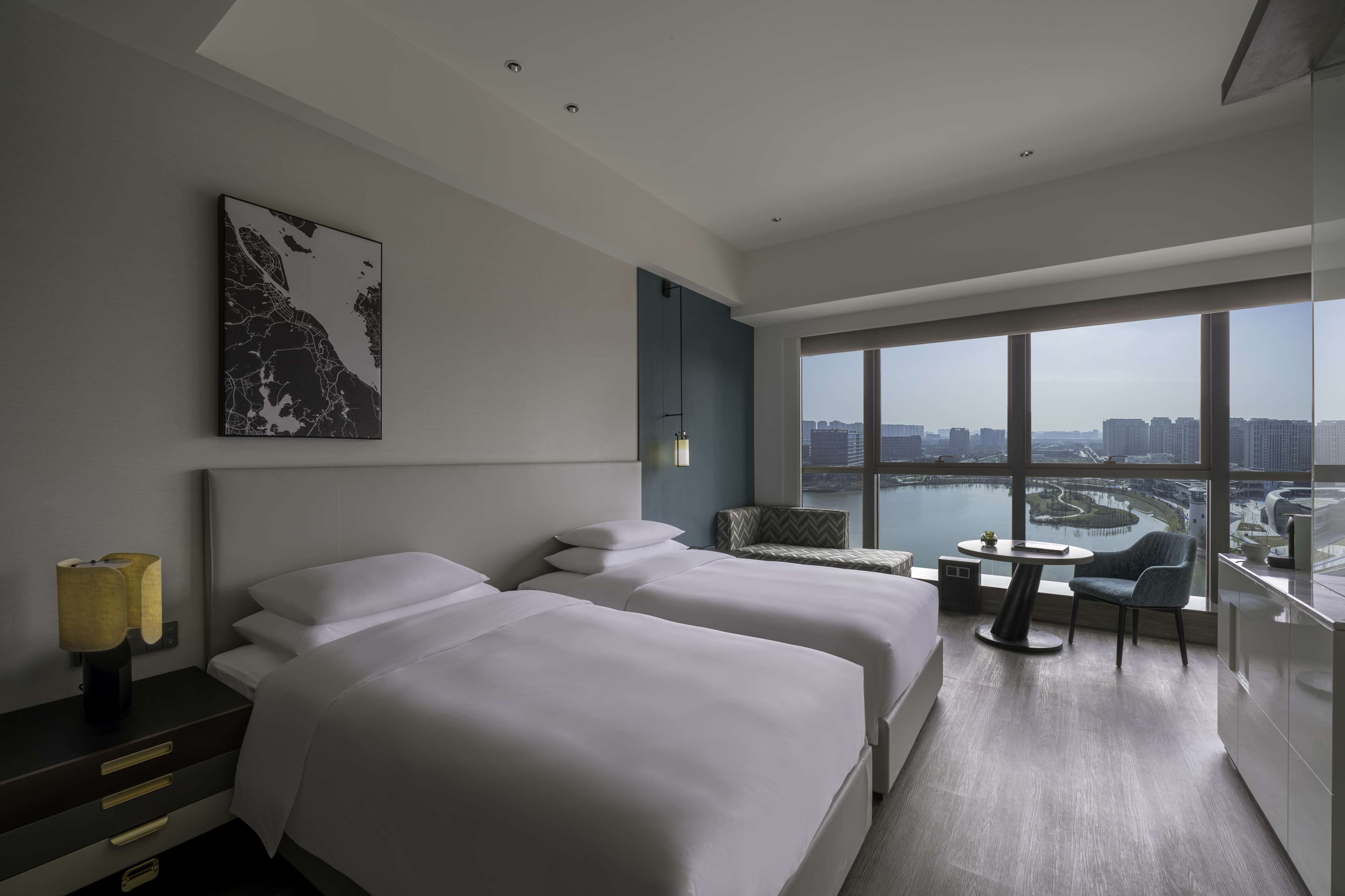 Quarto de hotel moderno com duas camas, um espaço de refeições e uma janela grande com vista para a cidade.