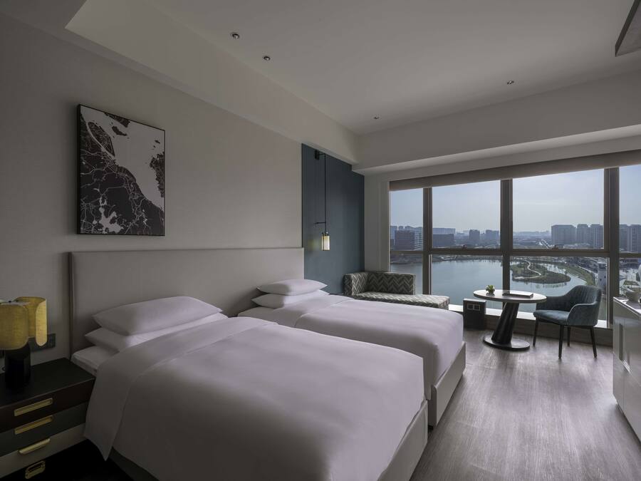 Quarto de hotel moderno com duas camas, um espaço de refeições e uma janela grande com vista para a cidade.