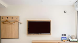 Quarto com guarda-roupa de madeira, escrivaninha com cadeira, televisão montada na parede e um banquinho.