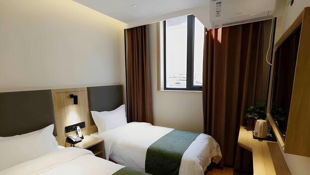 Quarto de hotel com duas camas, uma mesa de cabeceira com abajur, uma janela com cortinas e uma pequena mesa com uma planta.