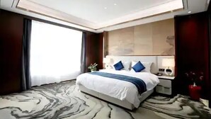 Quarto de hotel moderno com uma cama grande, um tapete com padrão marmorizado, uma mesa de cabeceira com abajur e vista para a cidade.