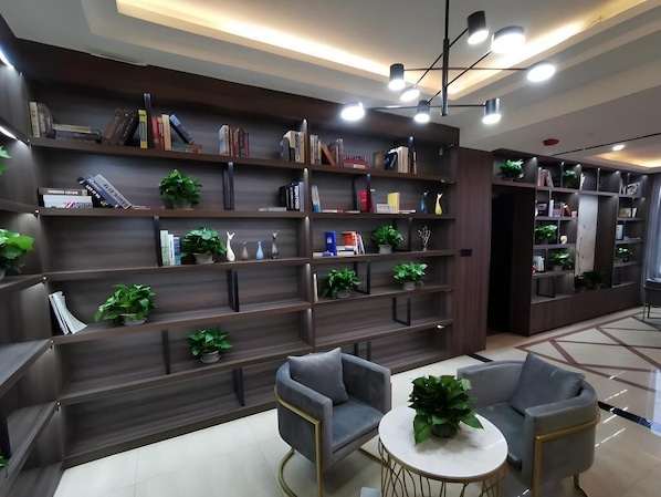 Uma biblioteca moderna com estantes de madeira escura, vários níveis e plantas verdes. Há duas poltronas cinzentas e uma pequena mesa redonda com uma planta.