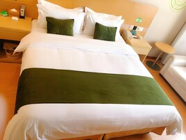 Uma cama bem arrumada com roupa de cama branca e verde, uma mesa de cabeceira com abajur e uma mesinha redonda com uma cadeira.