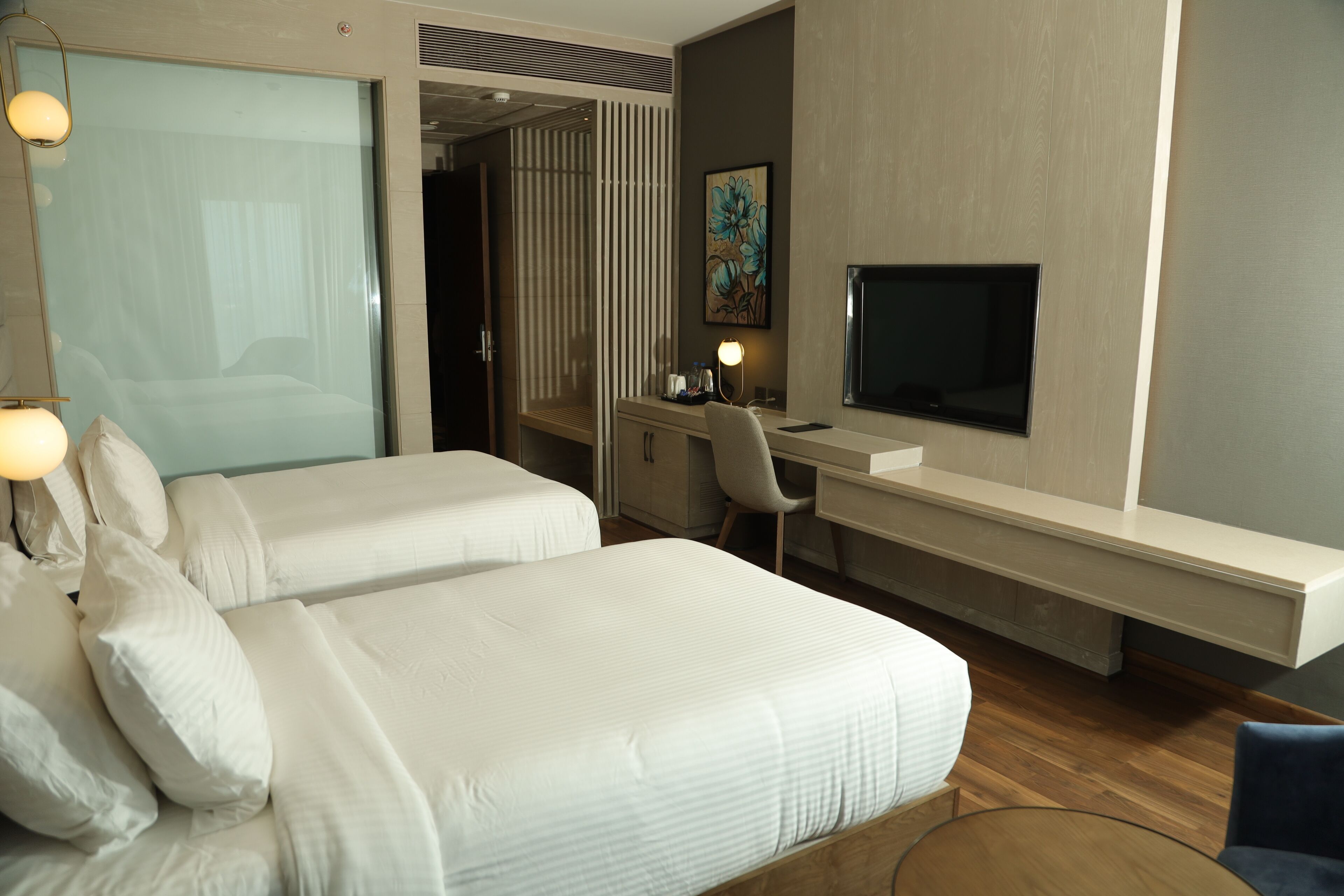 Quarto de hotel com duas camas, uma escrivaninha, uma cadeira, uma televisão e um quadro na parede.