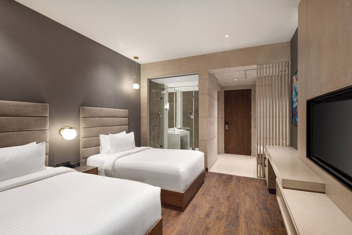 Quarto de hotel moderno com duas camas, televisão e banheiro com box de vidro.
