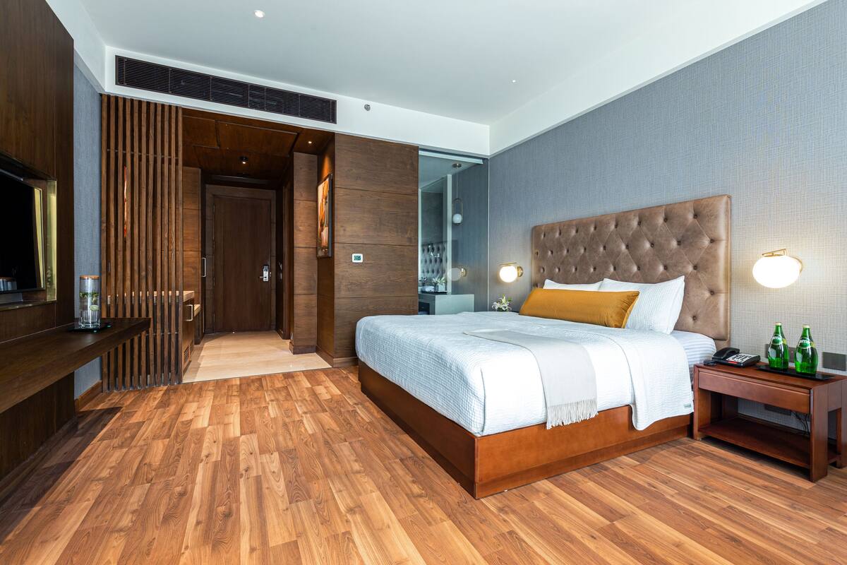 Quarto de hotel moderno com piso de madeira, uma cama grande com cabeceira estofada, uma mesa de cabeceira com abajur e painel de parede em madeira.