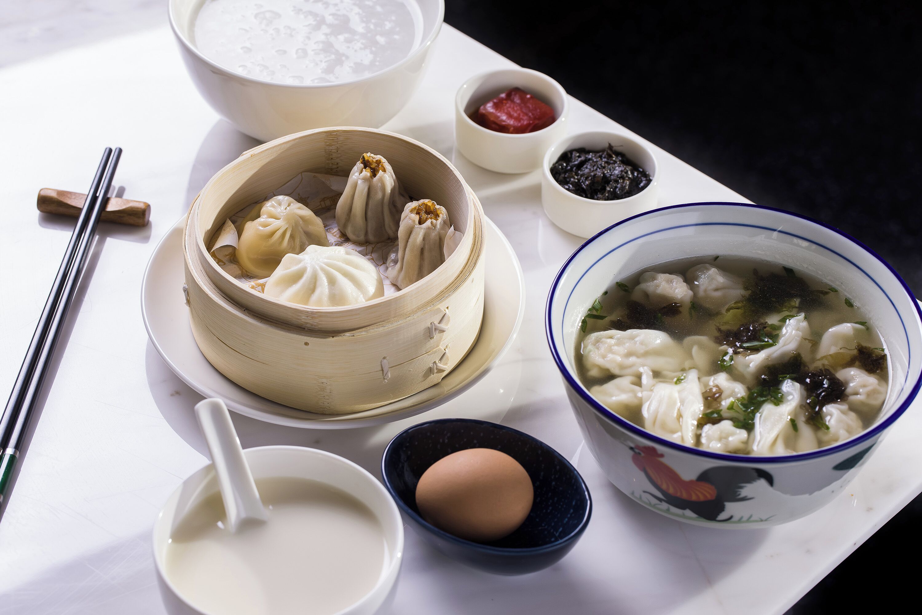 Uma mesa posta com uma tigela de sopa, um prato com dumplings, uma tigela de arroz, uma tigela de sopa e uma colher, uma tigela de molho de soja e uma tigela de ovo.