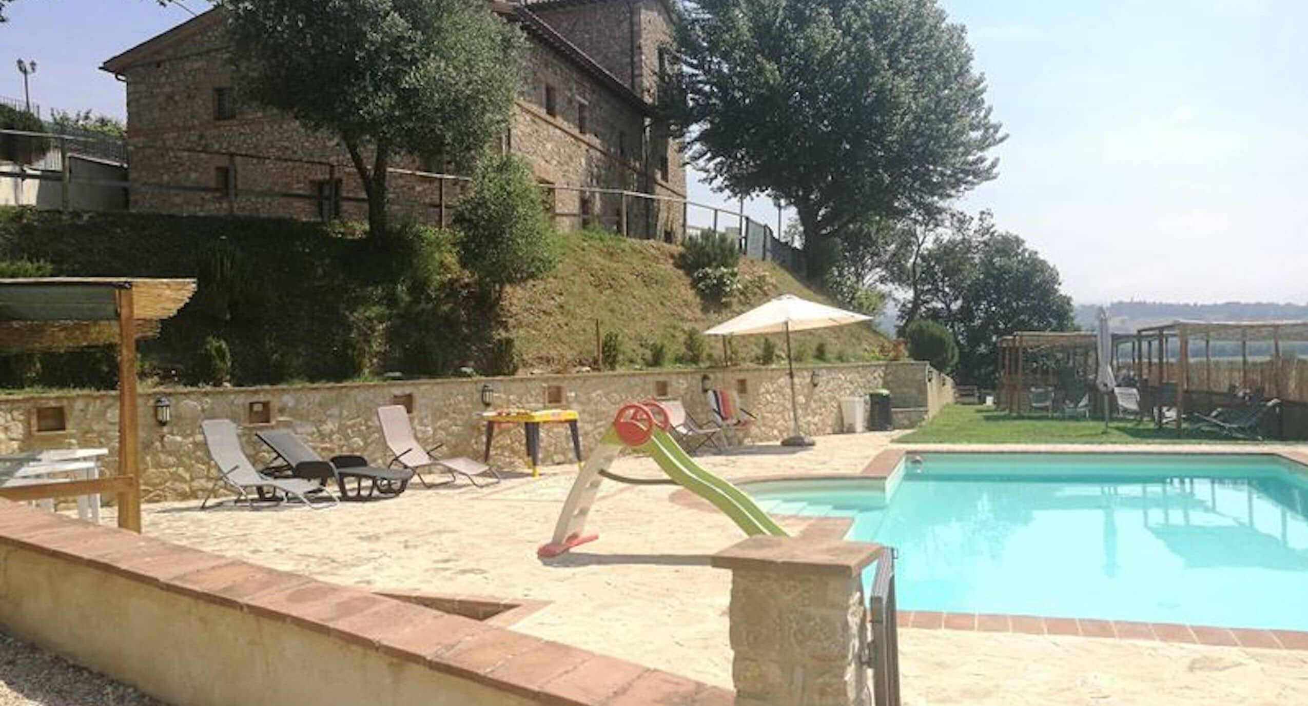 Uma área de piscina com espreguiçadeiras e um escorregador, rodeada por edifícios de pedra e árvores.