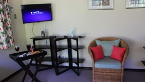 Quarto com televisão de tela plana exibindo 'DSTV', uma cadeira de vime com almofadas vermelhas, uma mesa pequena com uma bandeja contendo uma taça de vinho e um quadro na parede.