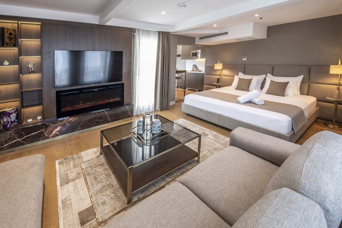 Quarto de hotel moderno com uma grande televisão de tela plana, um sofá, uma cama com roupa de cama branca e uma pequena cozinha.