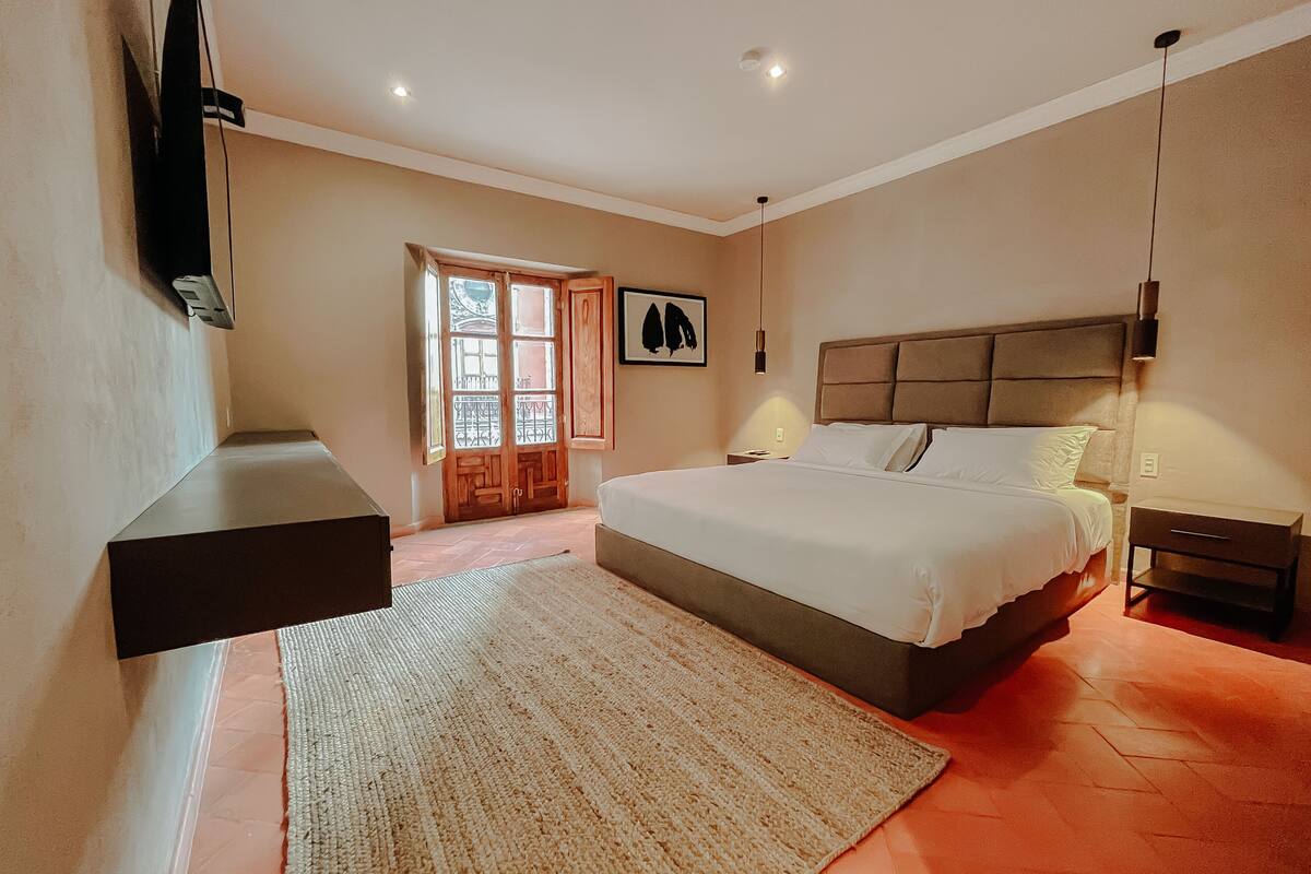 Um quarto com cama, mesas de cabeceira, televisão fixada na parede, porta com janela e tapete no chão.